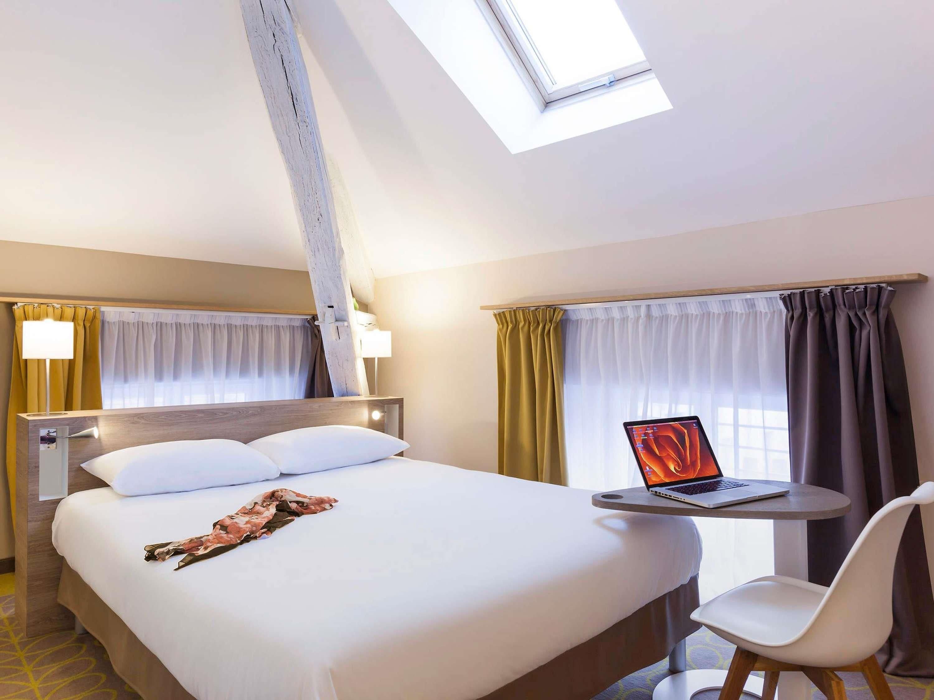 Quarto de hotel com uma cama, uma escrivaninha com um laptop, uma cadeira e um claraboia.