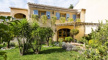 Um edifício em estilo mediterrâneo com um pátio, apresentando um arco central, uma varanda com persianas azuis e vegetação exuberante.
