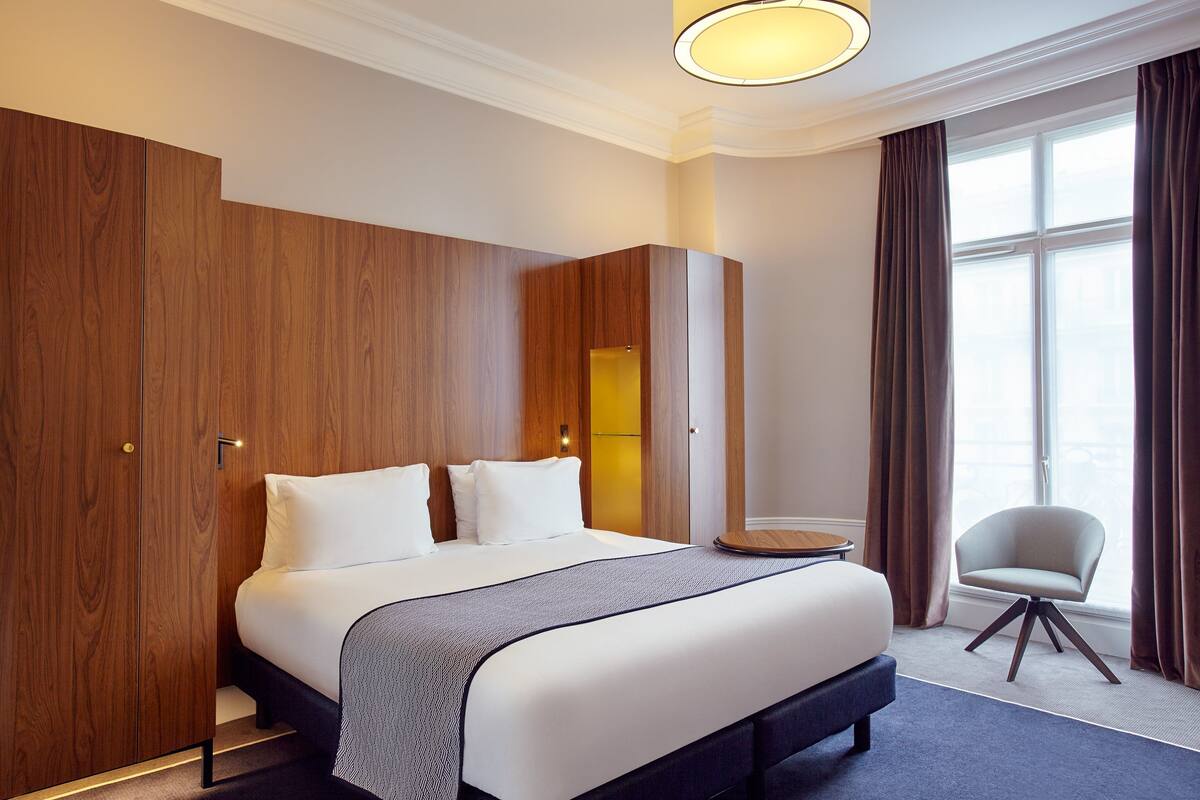 Quarto de hotel com uma cama grande, guarda-roupa de madeira, uma mesinha redonda, uma cadeira e uma janela com cortinas.
