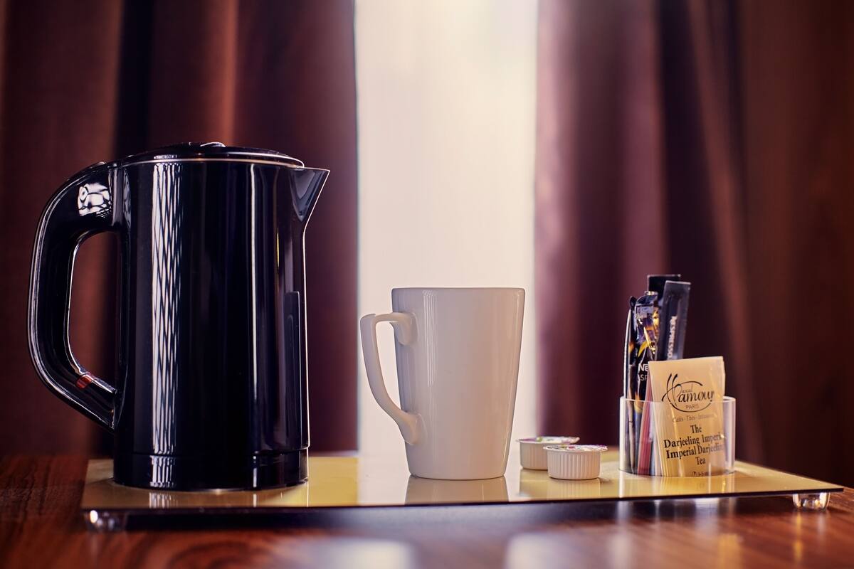 Uma chaleira elétrica preta, uma caneca branca e um porta-saquinhos de chá com sachês sobre uma bandeja espelhada.