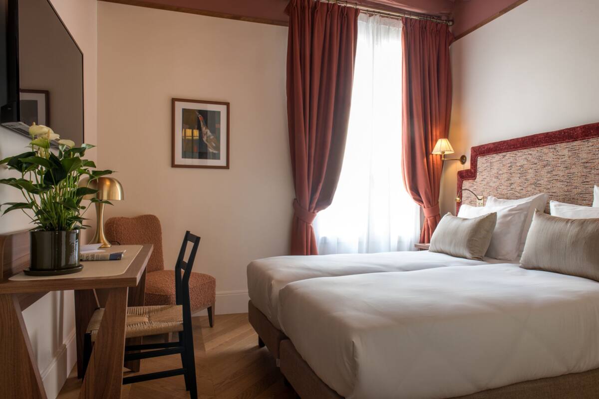 Quarto de hotel com cama, escrivaninha com cadeira, planta em vaso e um quadro na parede.