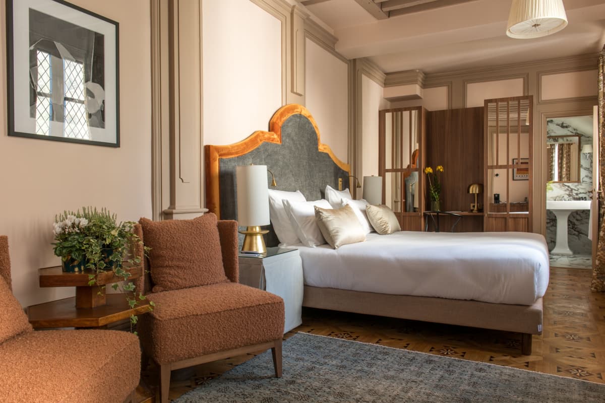 Quarto com uma cama grande, mesa de cabeceira de madeira, poltrona marrom e um espelho.