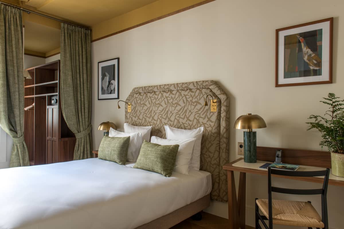 Quarto de hotel com cama, escrivaninha, cadeira, luminária, quadro emoldurado e planta em vaso.