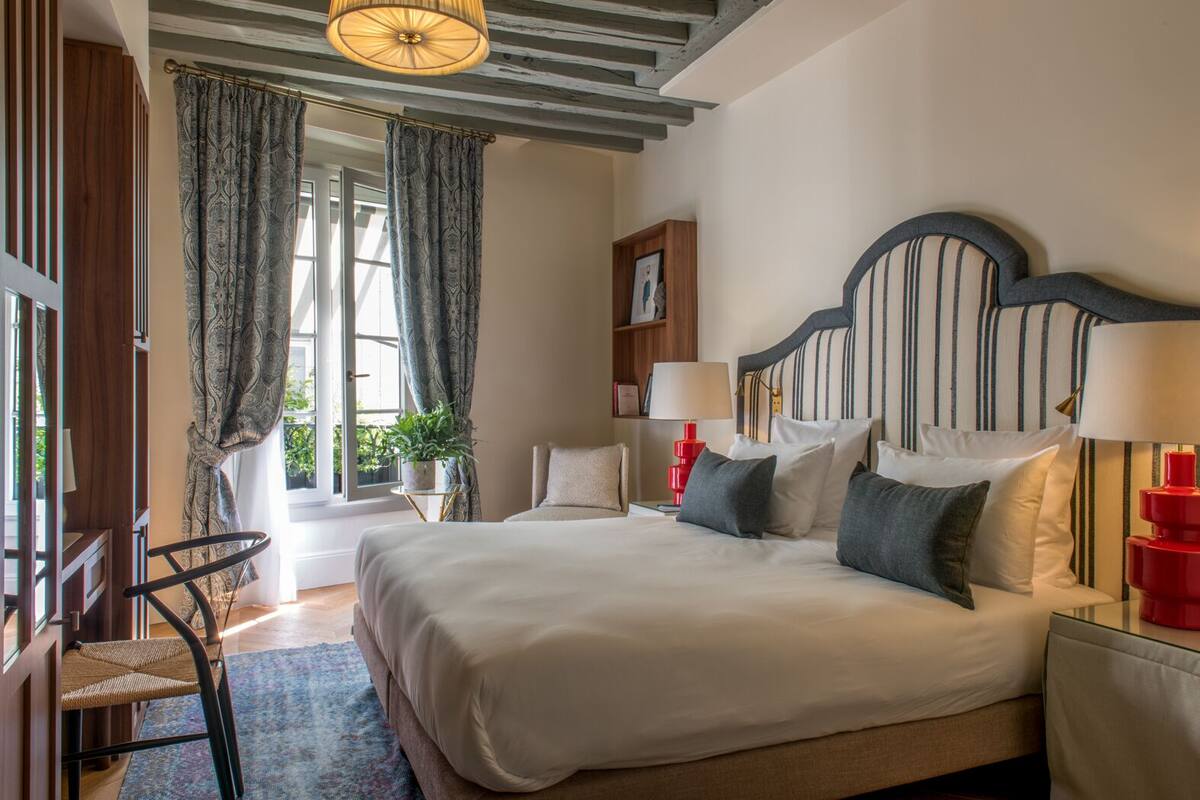 Quarto de hotel com uma cama grande, duas mesas de cabeceira com abajures, uma janela com cortinas, uma pequena mesa com uma planta e uma cadeira.