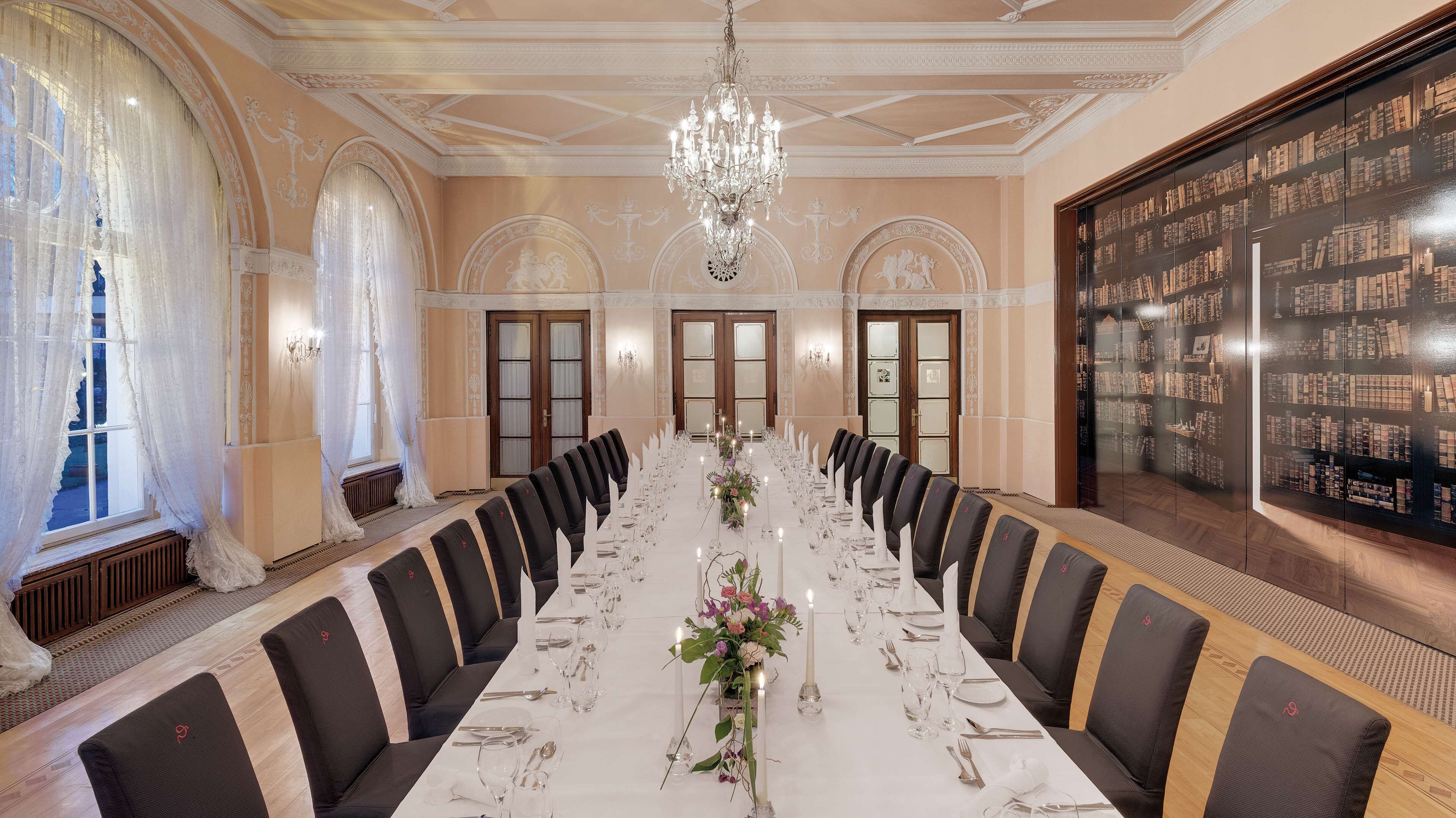 Uma sala de jantar formal com uma longa mesa posta para um banquete, ladeada por cadeiras pretas, um candelabro e uma estante com livros.