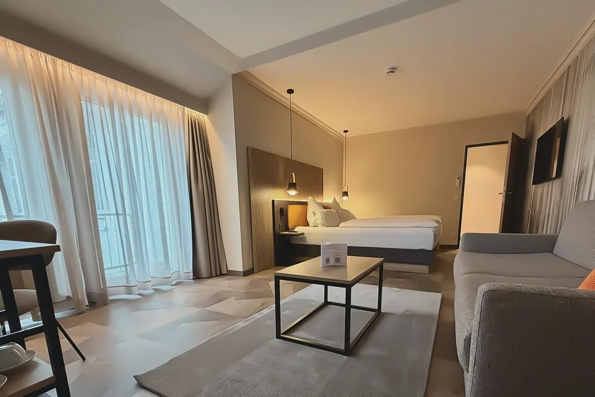 Quarto de hotel com cama, escrivaninha, cadeira, mesa de centro e uma grande janela com cortinas.