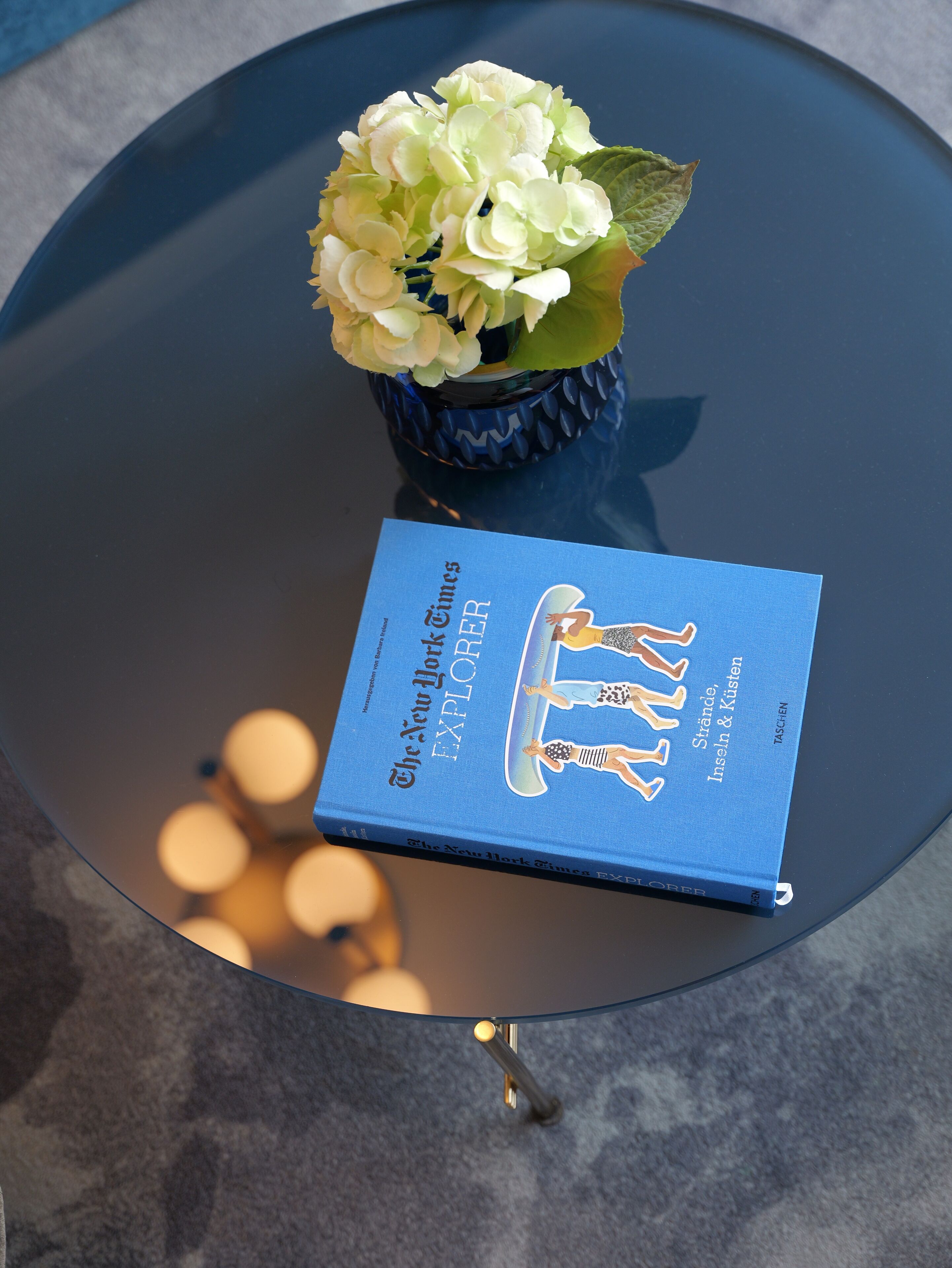 Um livro intitulado "The New York Times Explorer" sobre uma mesa de vidro com um vaso azul contendo flores brancas.