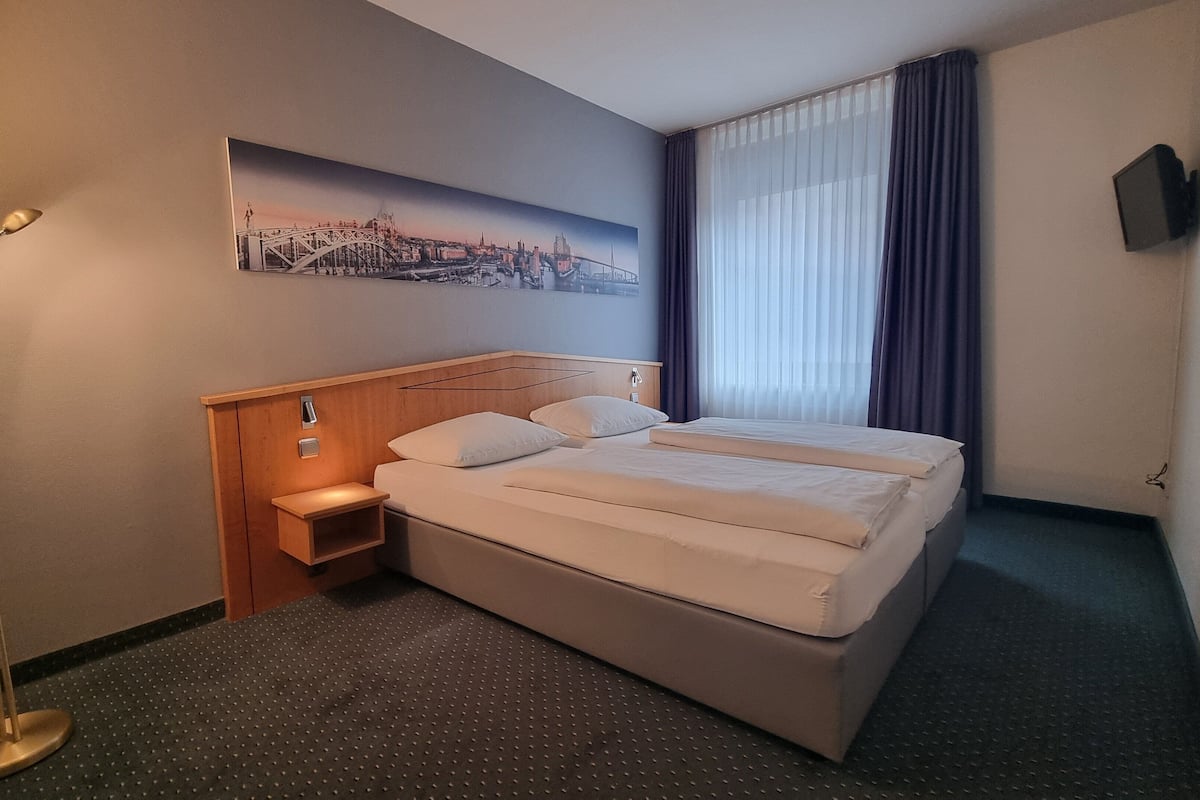 Quarto de hotel com cama, mesa de cabeceira, abajur, quadro e televisão na parede.