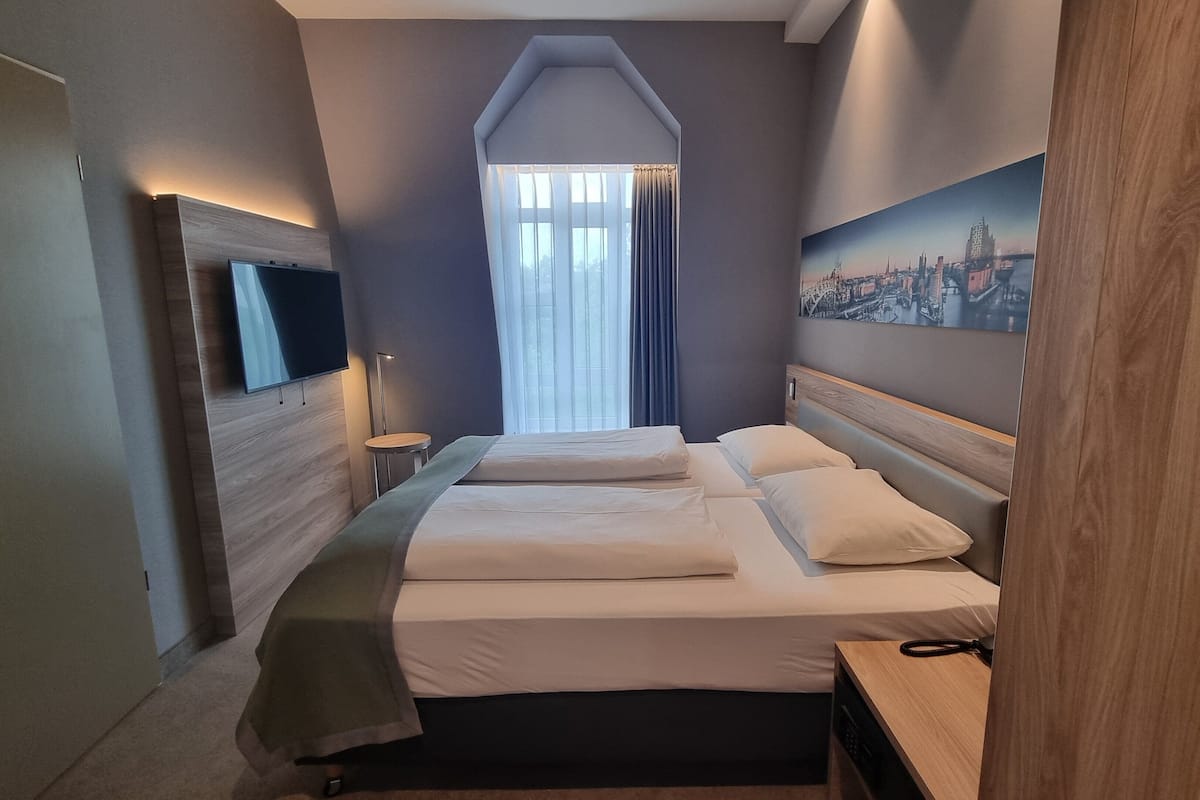 Quarto de hotel com cama, TV de tela plana, janela com cortinas e pintura de paisagem urbana na parede.