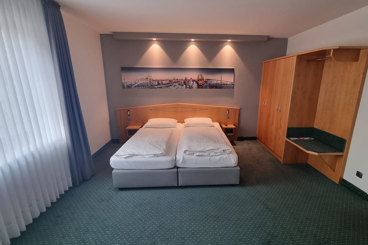 Quarto de hotel com cama de casal, armário de madeira, um pequeno espaço para sentar e uma obra de arte com vista para a cidade na parede.