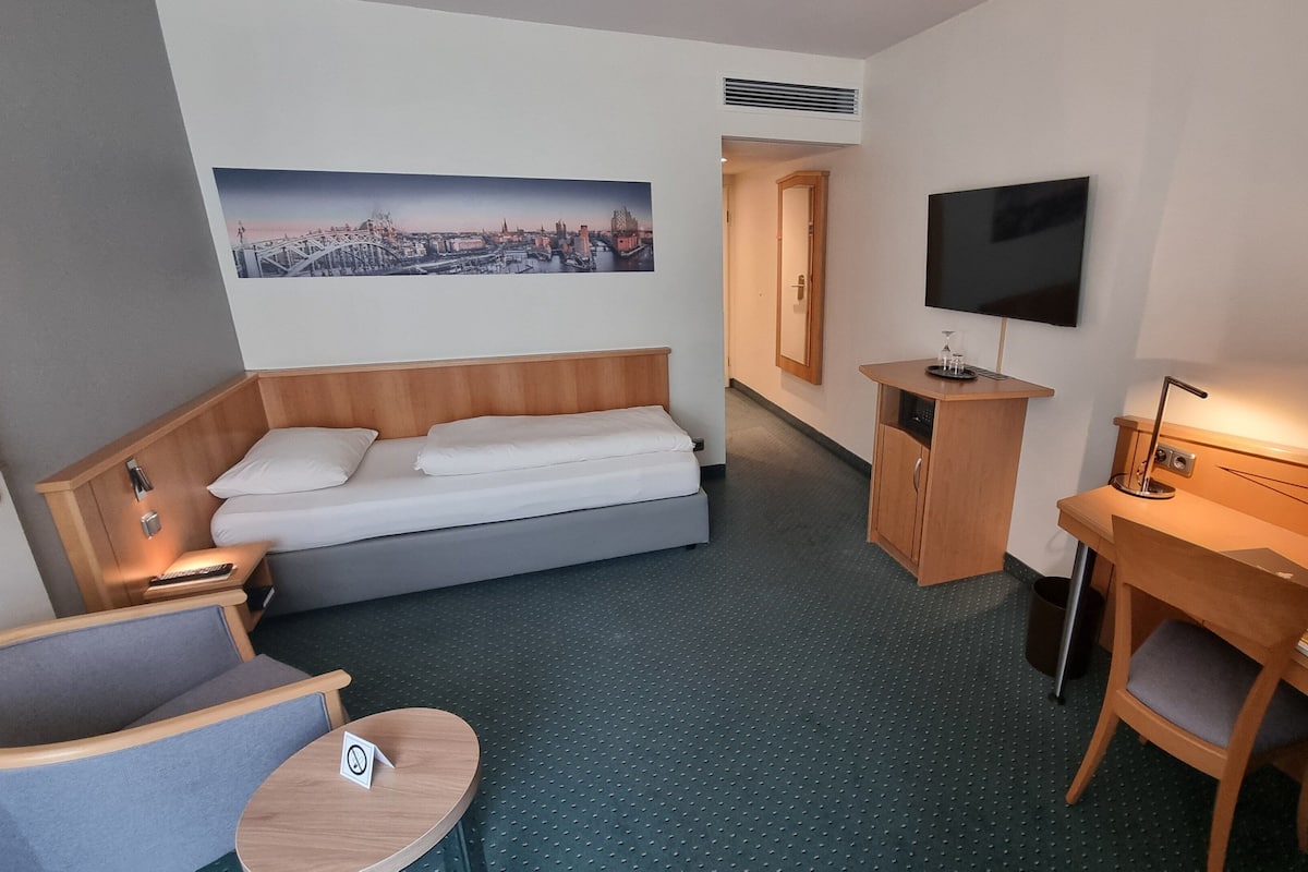 Quarto de hotel com cama, escrivaninha, cadeira, televisão e pintura de uma paisagem urbana.