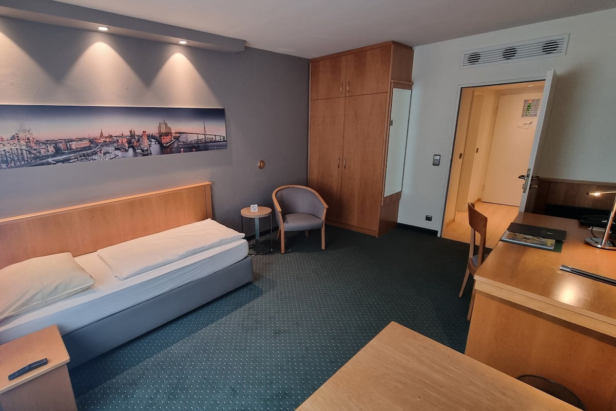 Quarto de hotel com cama, mesa redonda pequena, cadeira, guarda-roupa de madeira e um quadro pendurado na parede.
