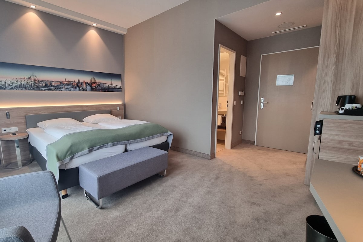 Quarto de hotel moderno com uma cama, mesa de cabeceira e uma grande pintura de paisagem urbana na parede.