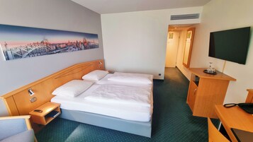 Quarto de hotel com cama, escrivaninha, cadeira, televisão e um quadro na parede.