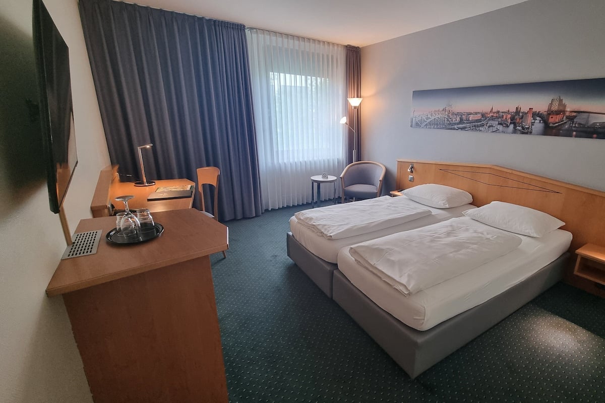 Quarto de hotel com duas camas, uma escrivaninha, uma cadeira, uma mesinha e um quadro na parede.