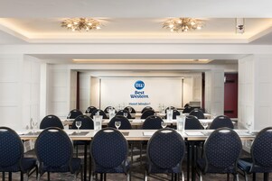Uma sala de conferências com uma mesa comprida, cadeiras e uma tela exibindo o logotipo do Hotel Best Western Windorf.