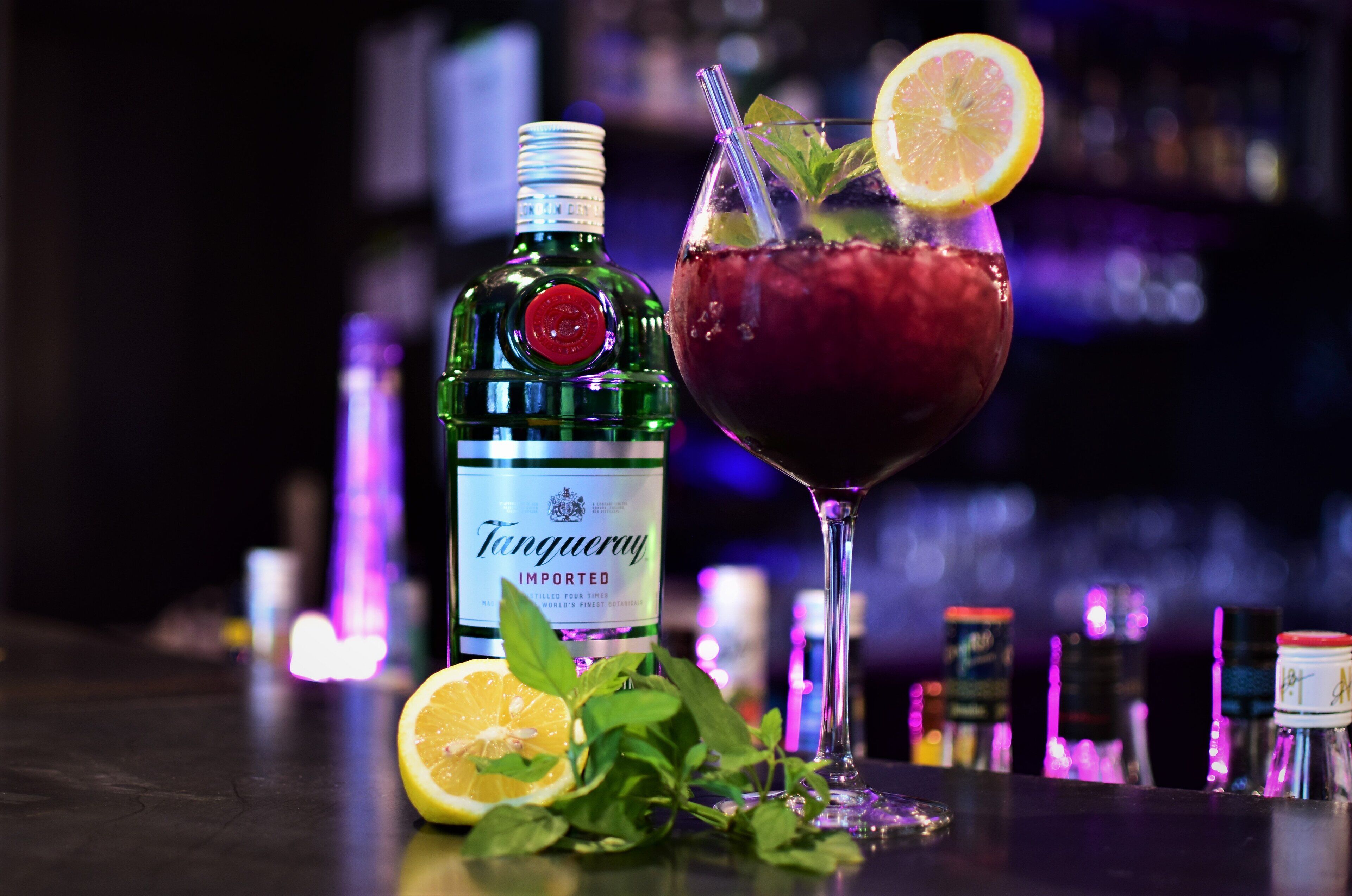 Uma garrafa de licor Tanguray Imported, um copo de cocktail vermelho com uma fatia de limão e hortelã, e meia lima em um balcão de bar.