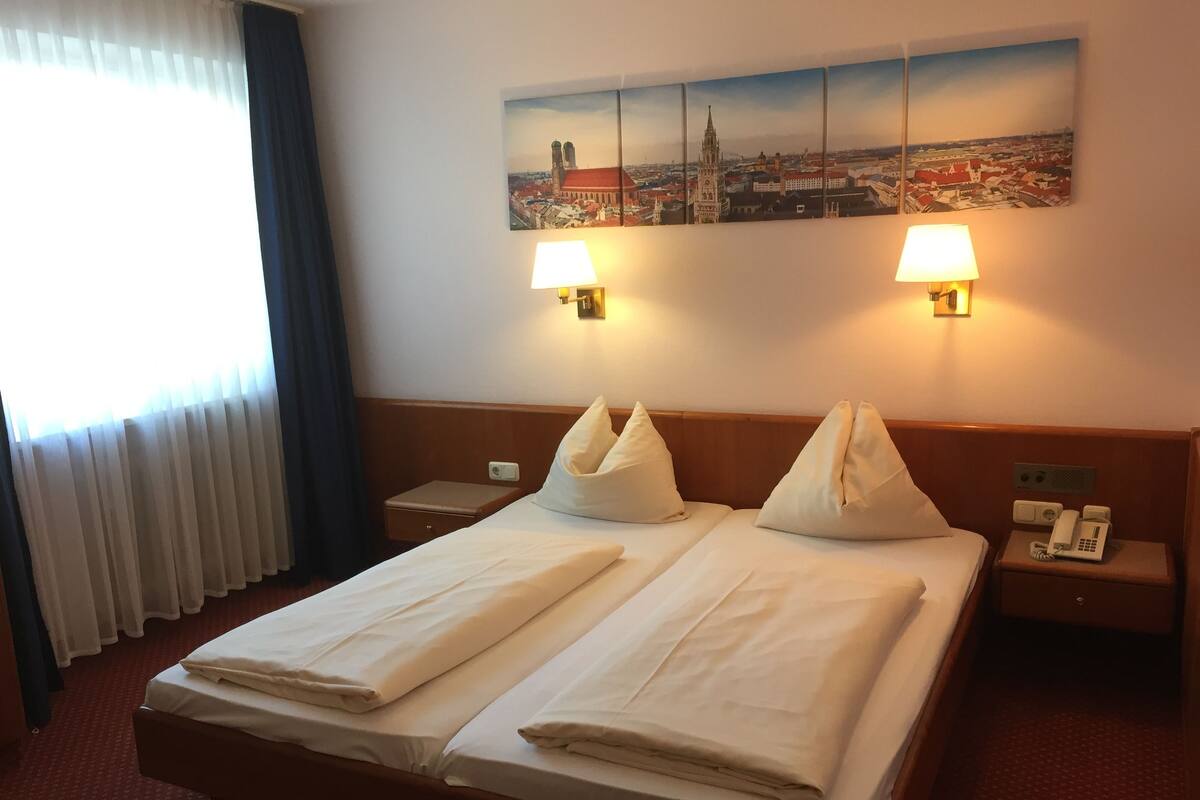 Quarto de hotel com uma cama grande, duas mesas de cabeceira, um criado-mudo com telefone e um quadro com vista para a cidade na parede.