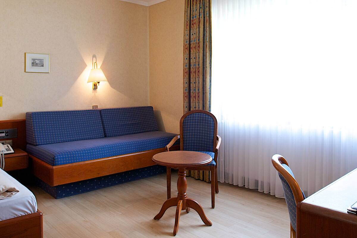 Quarto de hotel com cama, sofá, poltrona, escrivaninha, televisão e duas luminárias fixadas na parede.