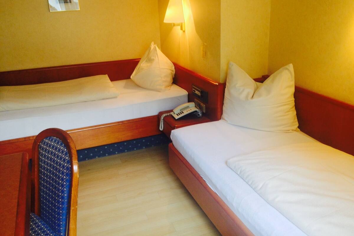 Quarto de hotel com duas camas de solteiro, uma cadeira de madeira, uma mesa de cabeceira com luminária e um telefone.