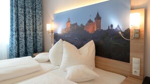 Quarto de hotel com uma cama grande, duas luminárias fixadas na parede e um painel mural com vista para um castelo.