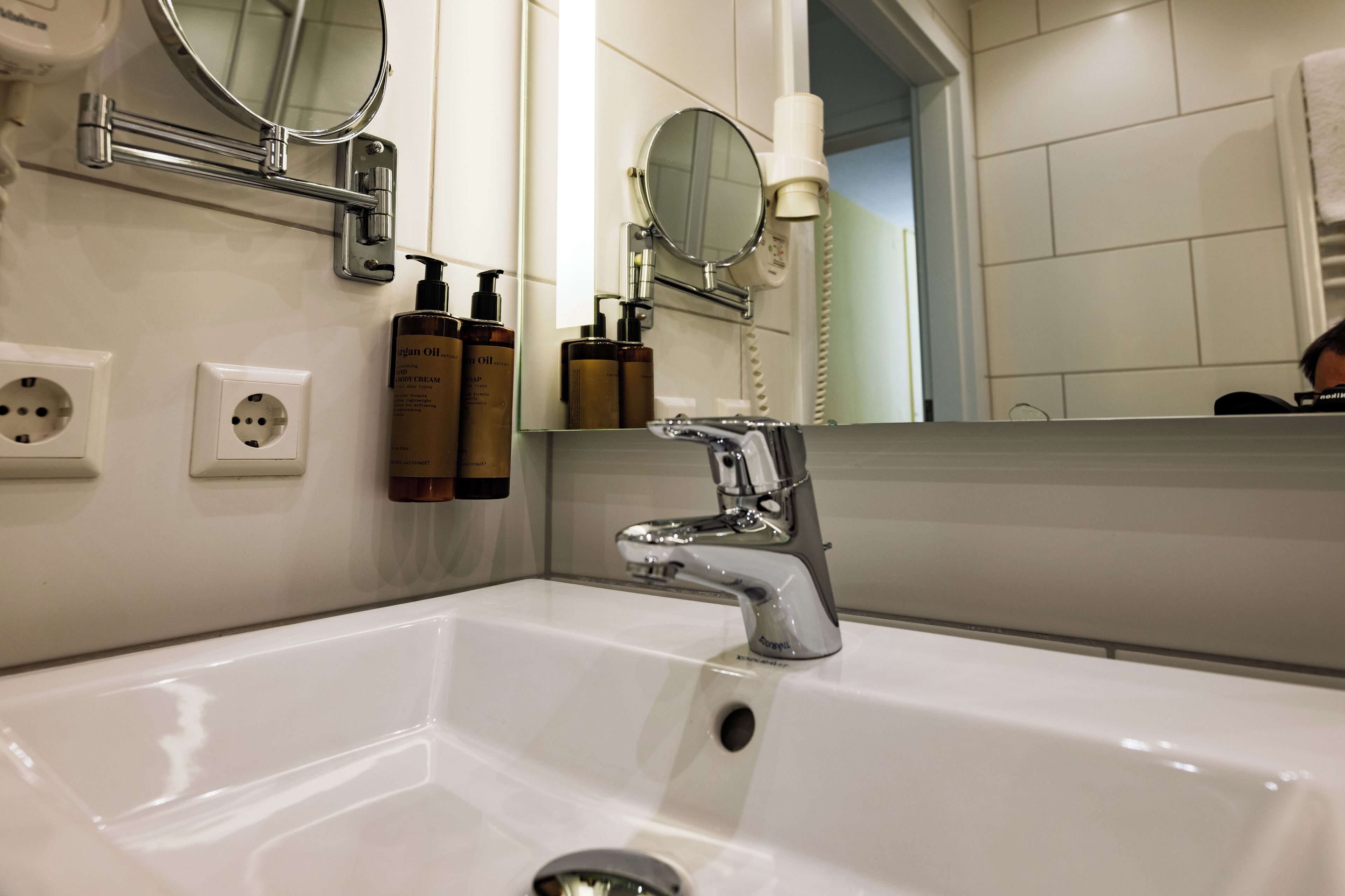 Casa de banho com lavatório branco, torneira cromada e duas espelhas. Há produtos de higiene pessoal na parede e um secador de cabelo no balcão.