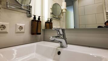 Casa de banho com lavatório branco, torneira cromada e duas espelhas. Há produtos de higiene pessoal na parede e um secador de cabelo no balcão.