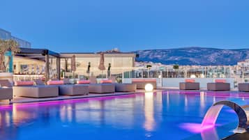 Uma área de piscina no terraço com assentos de lounge e vista para a paisagem urbana e montanhas.