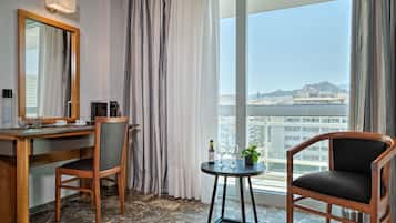 Quarto de hotel com escrivaninha, duas cadeiras, uma mesa pequena com uma garrafa e copos, um espelho e uma janela grande com vista para a cidade.