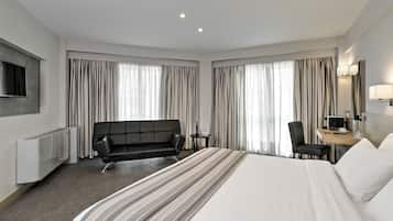 Quarto de hotel com uma cama grande, um sofá, uma escrivaninha com cadeira, uma televisão fixada na parede e uma janela com cortinas.