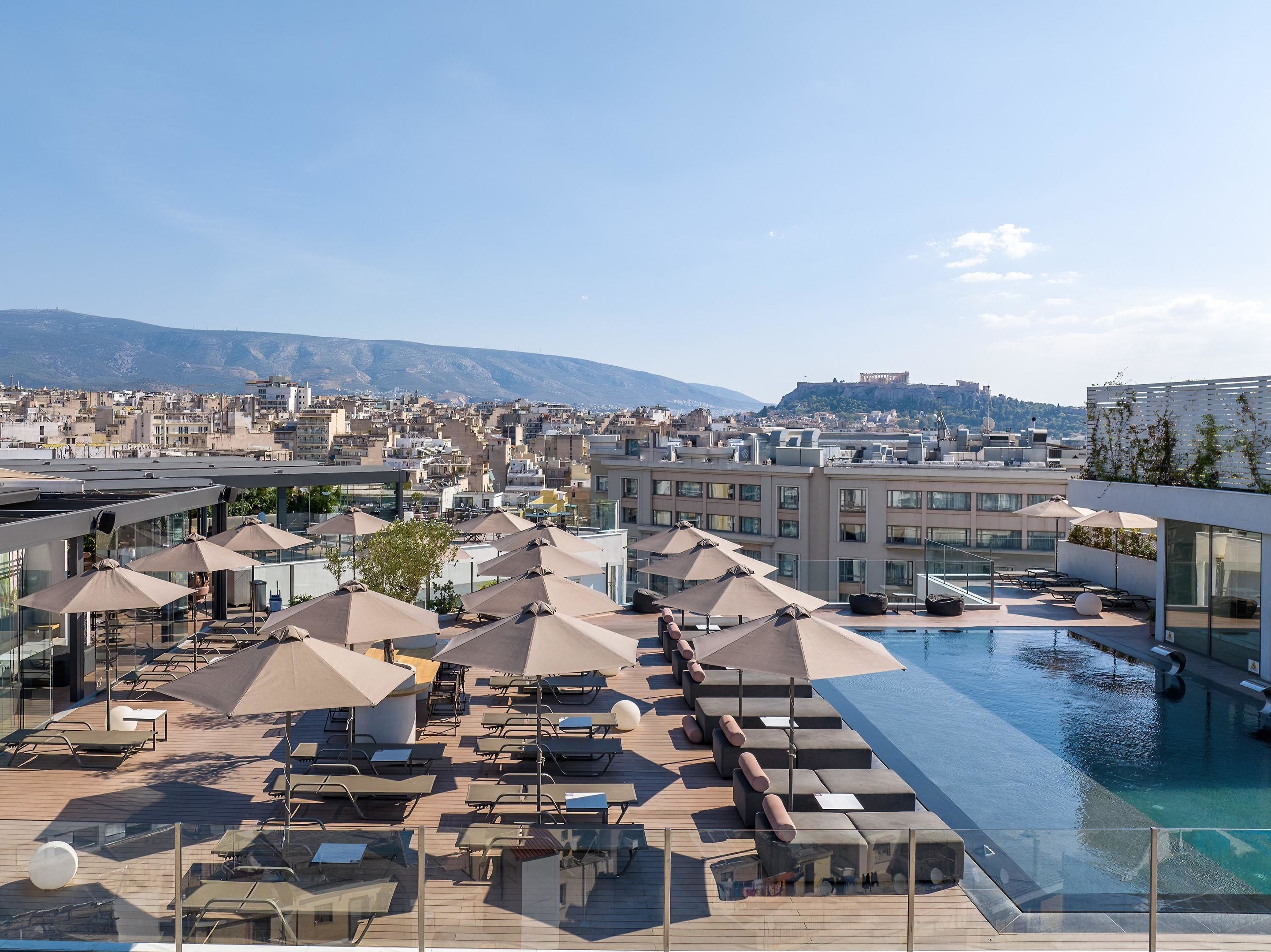 Área de piscina no terraço com espreguiçadeiras e guarda-sóis, com vista para uma paisagem urbana e montanhas ao fundo.