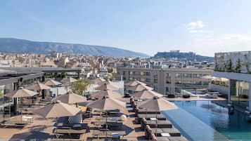 Área de piscina no terraço com espreguiçadeiras e guarda-sóis, com vista para uma paisagem urbana e montanhas ao fundo.