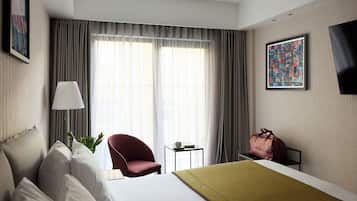 Quarto de hotel com cama, uma cadeira, uma mesinha de cabeceira com abajur, televisão e um quadro na parede.
