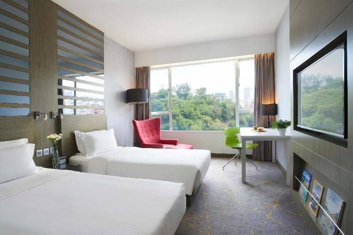 Quarto de hotel com duas camas, uma escrivaninha, uma cadeira, televisão e uma janela grande com vista para o verde.
