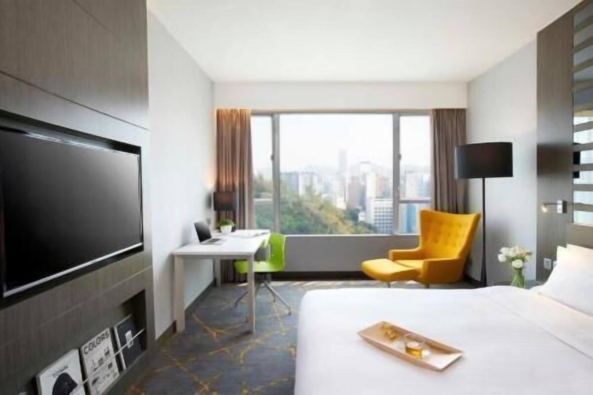 Quarto de hotel com cama, escrivaninha, cadeira, televisão e janela com vista para a cidade.