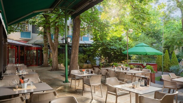 Um café ao ar livre com mesas e cadeiras, cercado por árvores e coberto por um toldo verde.