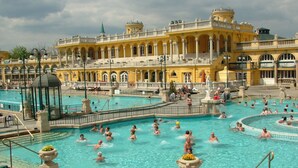 Uma grande piscina ao ar livre com várias áreas, rodeada por um edifício clássico com arcos e colunas.
