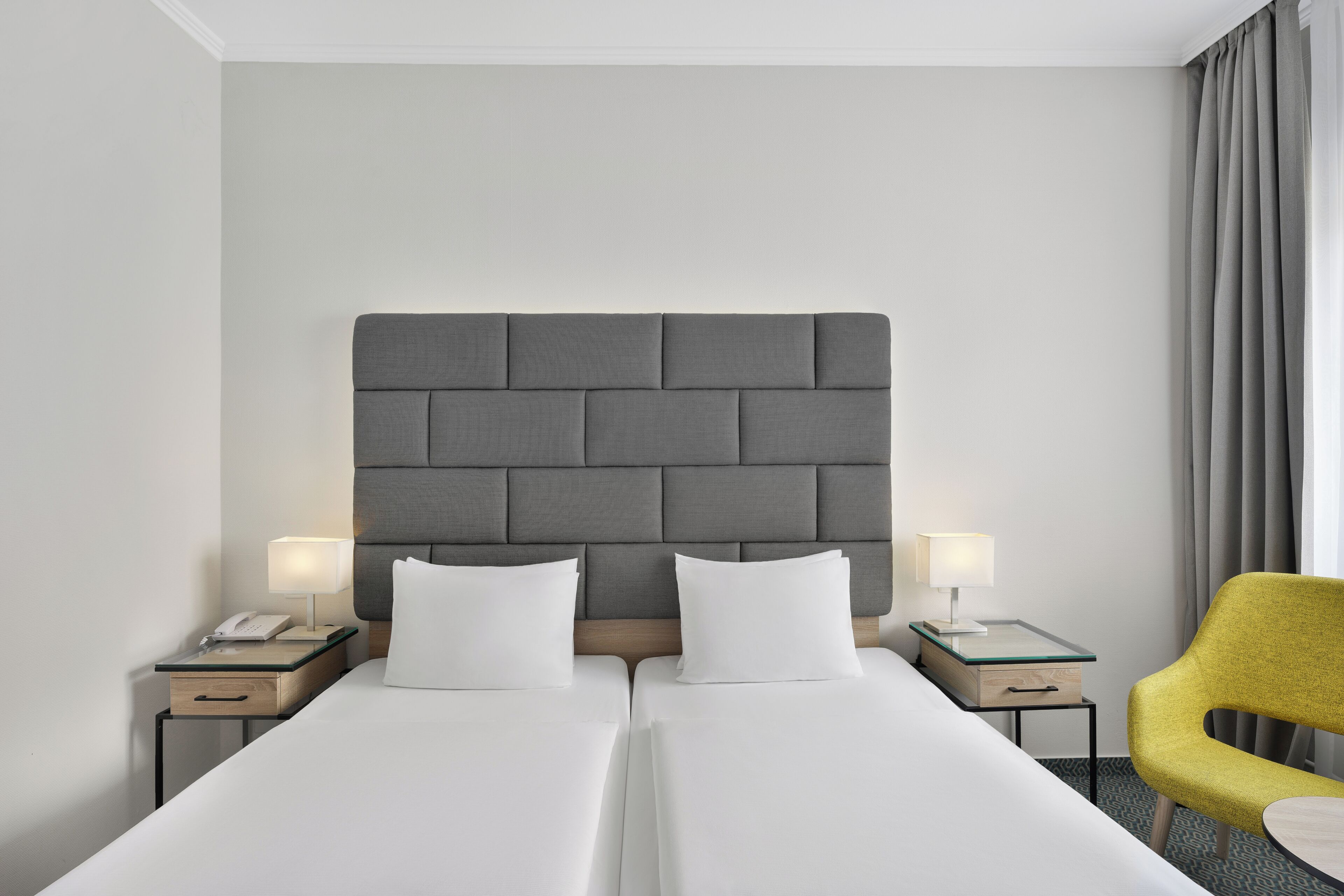 Quarto de hotel com duas camas, cabeceira estofada cinza, mesas de cabeceira com luminárias, uma cadeira amarela e uma pequena mesa.