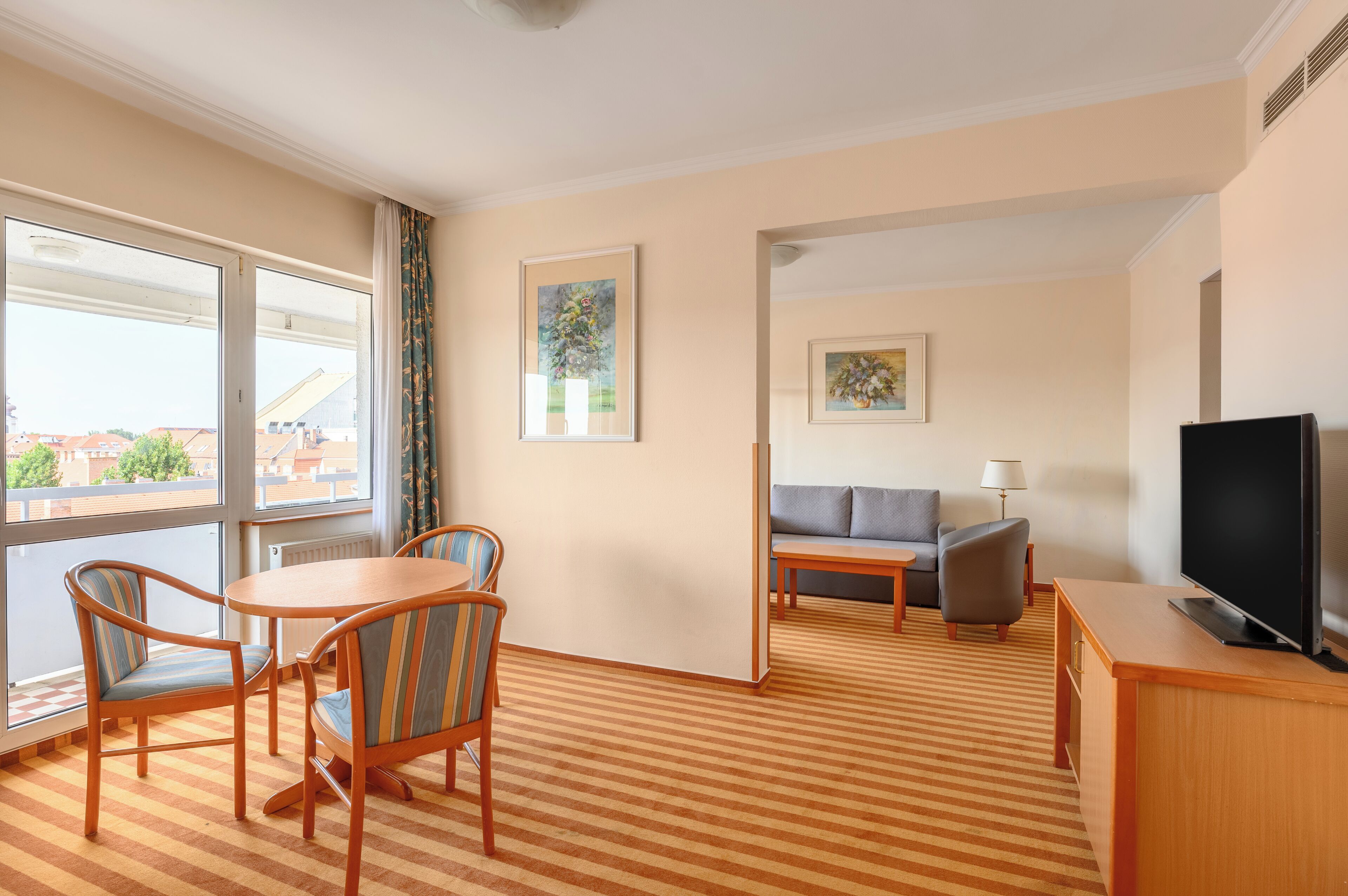 Quarto de hotel com área de refeições, sofá, televisão e janelas amplas.