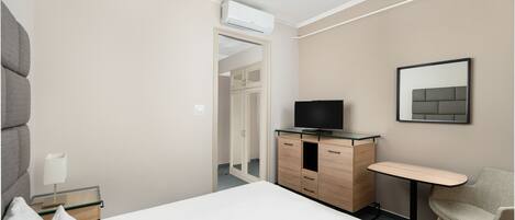 Quarto de hotel com uma cama, uma televisão sobre um móvel de madeira, uma escrivaninha, uma cadeira e um guarda-roupa.