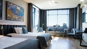 Quarto de hotel com duas camas, um sofá, uma escrivaninha e uma janela grande com vista para a cidade.