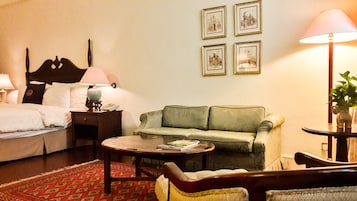 Quarto de hotel com cama, sofá verde, mesa pequena, luminária e quadros na parede.
