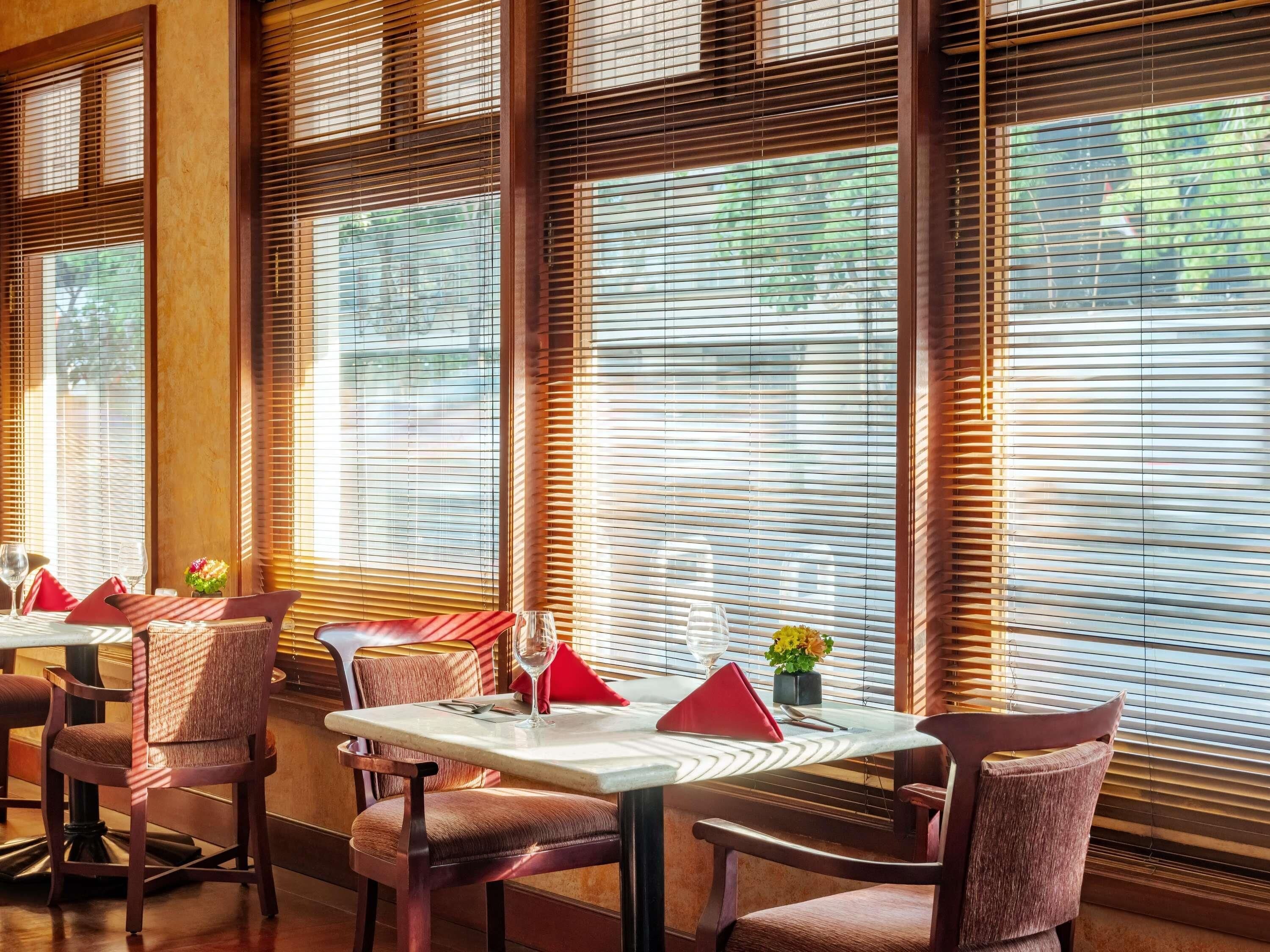 Interior de restaurante com persianas de madeira, mesas redondas, cadeiras acolchoadas e pequenas plantas em vasos.