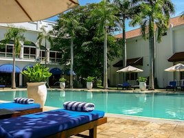 Área de piscina com espreguiçadeiras e guarda-sóis, cercada por palmeiras e um edifício com telhado de azulejos.