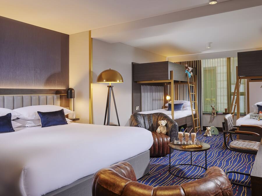 Quarto de hotel com duas camas, uma escrivaninha com abajur, uma cadeira, uma pequena mesa com bandeja e um urso de pelúcia em uma das camas.