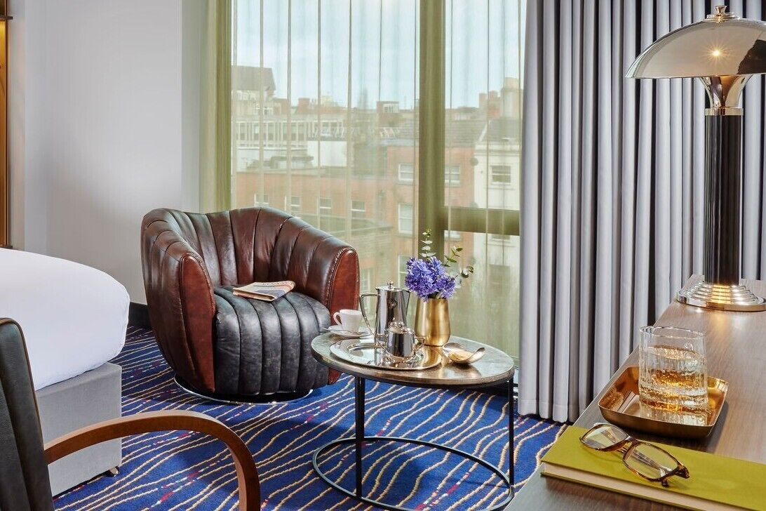 Quarto de hotel com poltrona de couro, uma mesa redonda pequena com bandeja, abajur e vista para prédios pela janela.