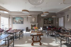 Uma sala de jantar ampla, com várias mesas e cadeiras, uma lareira central e grandes pinturas nas paredes.