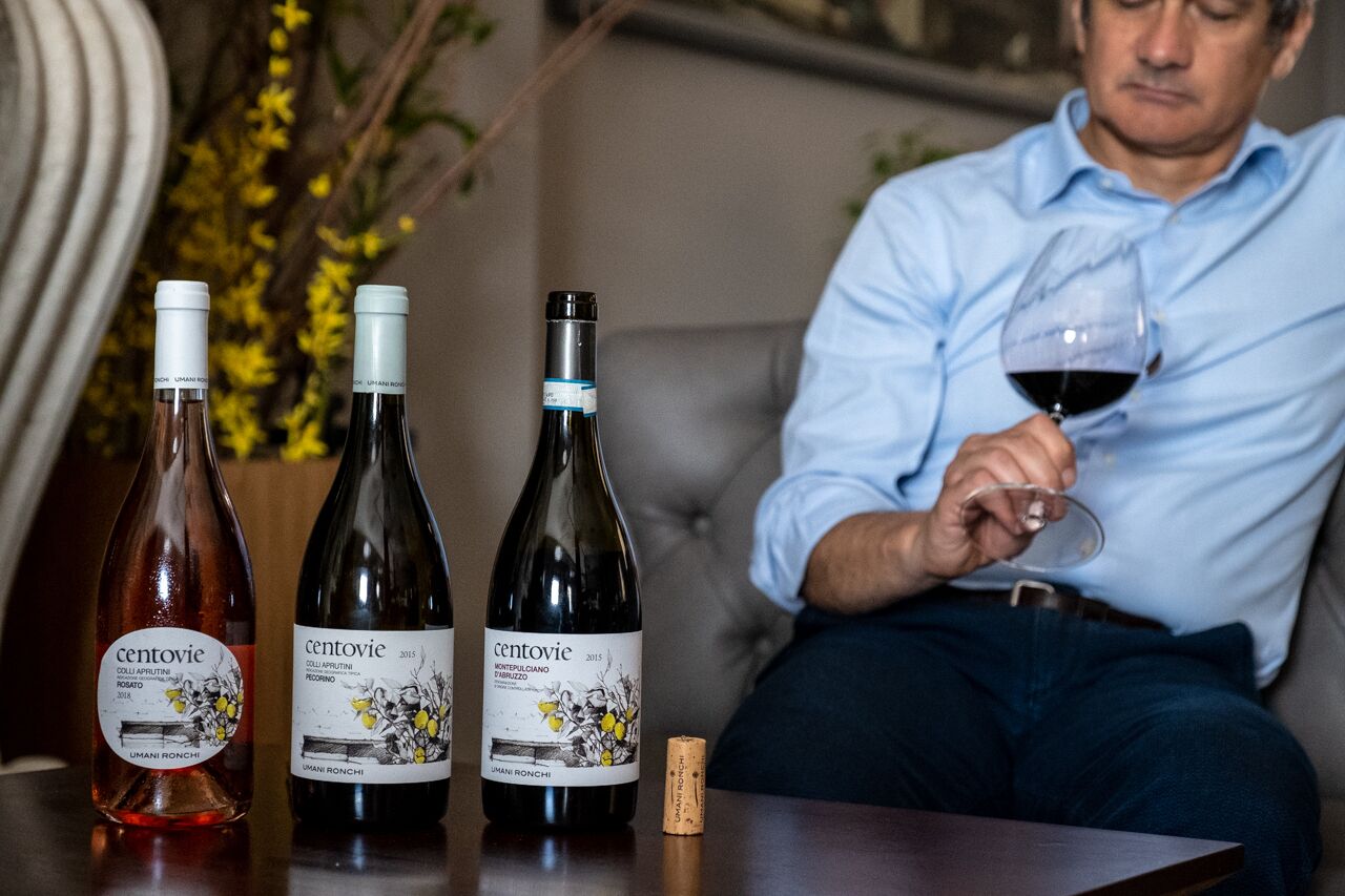 Três garrafas de vinho Centovie sobre uma mesa, com uma rolha na frente. Um homem segura uma taça de vinho.
