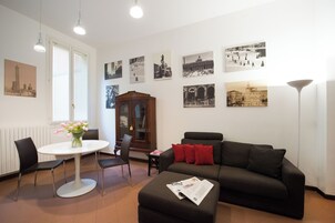 Uma sala de estar com um sofá preto, uma mesa de jantar redonda com cadeiras, um armário de madeira e um vaso de flores.