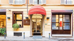A entrada do HOTEL PHI BOLOGNA com toldo vermelho, porta de entrada visível e uma vitrine com um manequim.